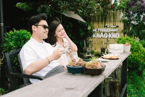 Um casal sentado em uma mesa ao ar livre, com um cenário de bambu e plantas em vasos.