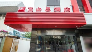 Entrada de hotel com fachada em vermelho e branco, portas de vidro e área de estar interna visível.