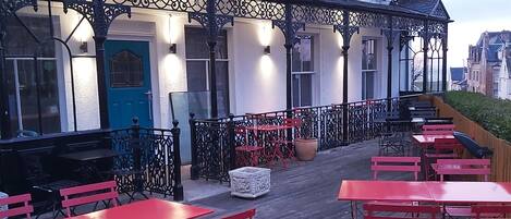 Um edifício com terraço, apresentando mesas e bancos vermelhos, uma porta azul e um par de chifres decorativos fixados na parede.