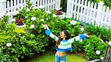 Uma pessoa em um jardim com cerca branca de piquete, rodeada por vegetação exuberante e flores brancas.