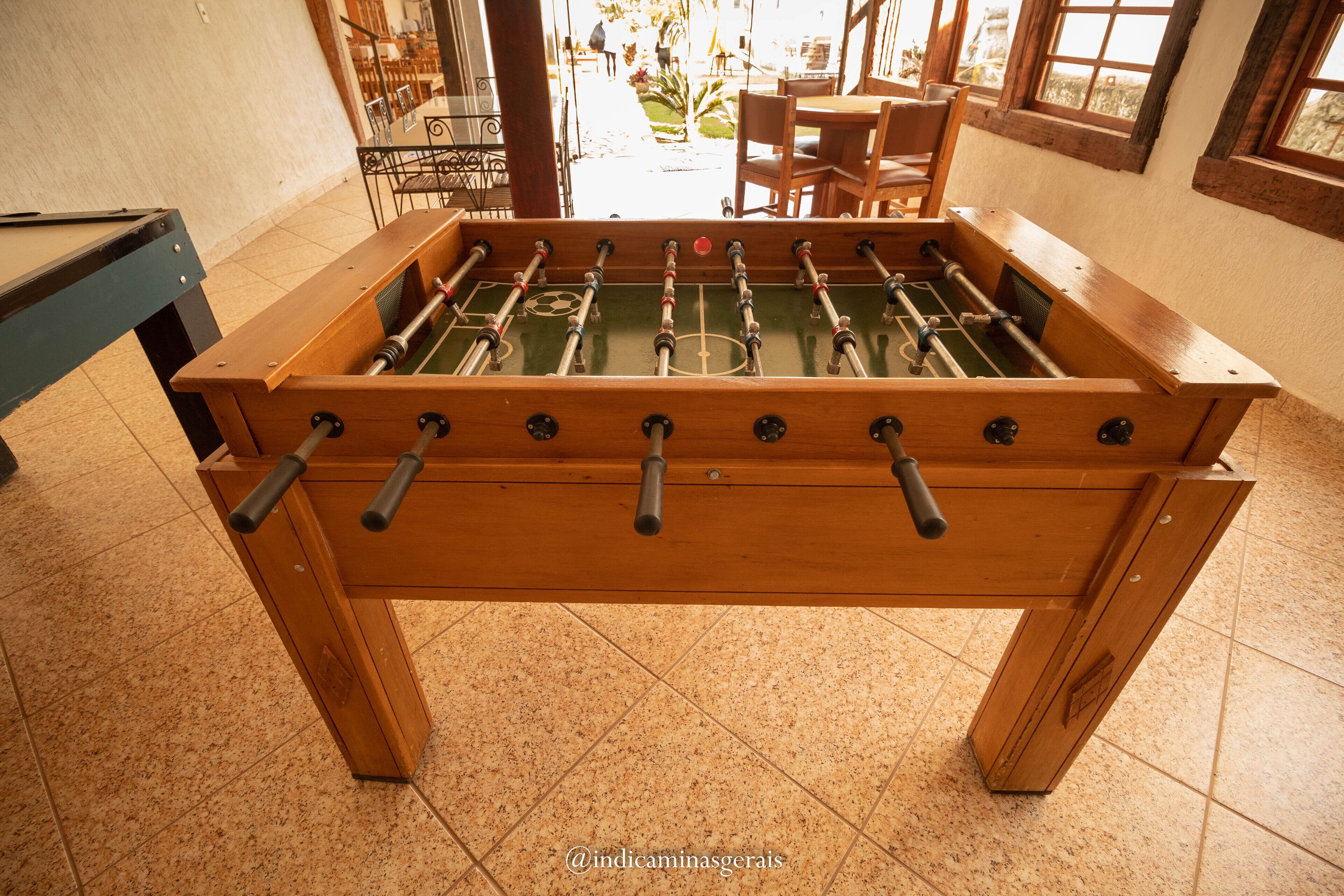 Uma mesa de futebol de madeira em um espaço com piso de azulejos e vista para área externa através de uma janela.
