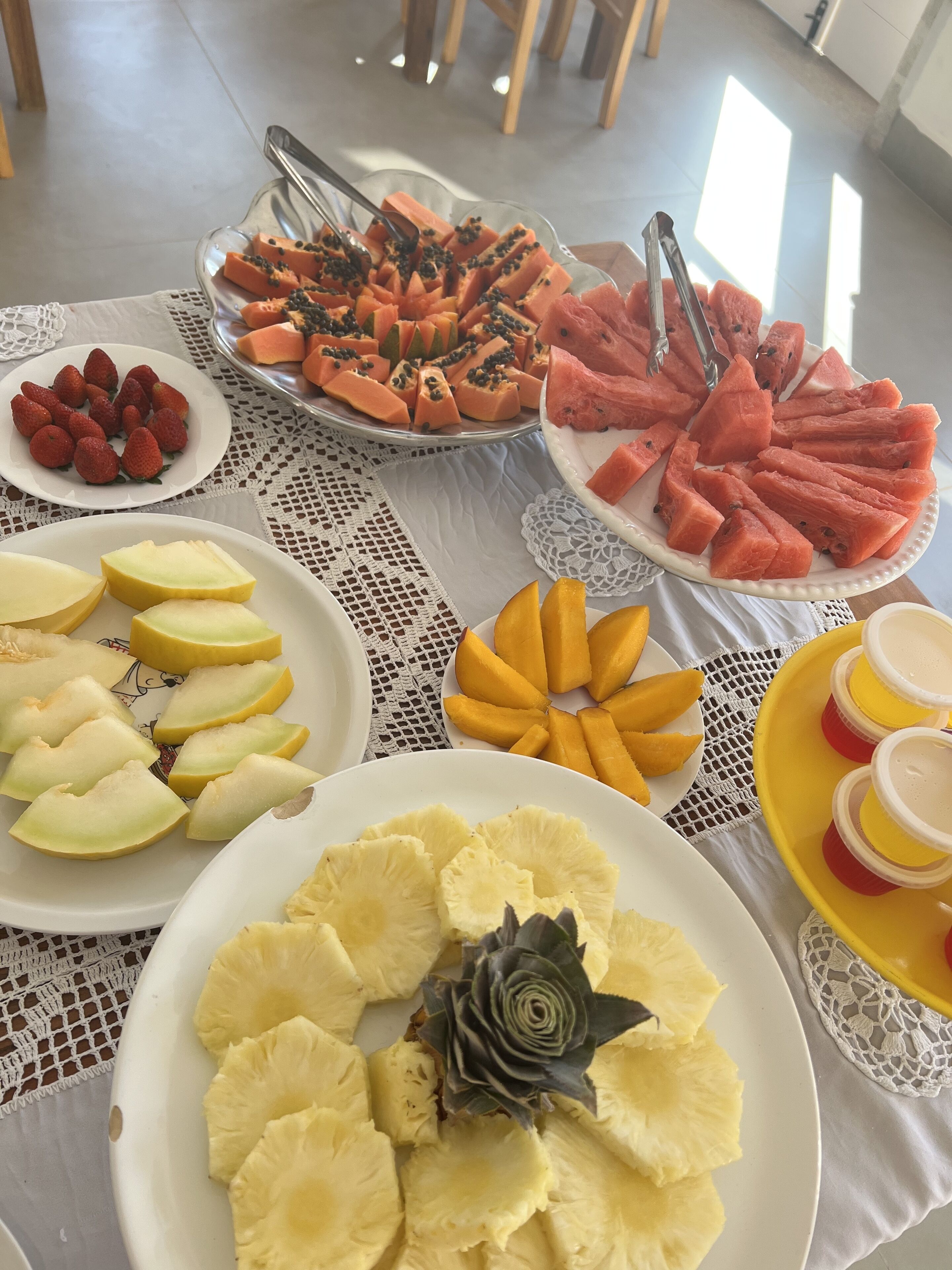 Uma mesa com diversos tabuleiros de frutas, incluindo mamão, melancia, morangos, melão e abacaxi em fatias.