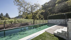 Piscina com deck de madeira, parede de pedra e vista para vinhedos e colinas.