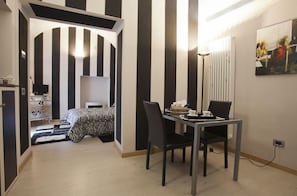Quarto com cama listrada em preto e branco, mesa de jantar com cadeiras, radiador branco e um quadro na parede.