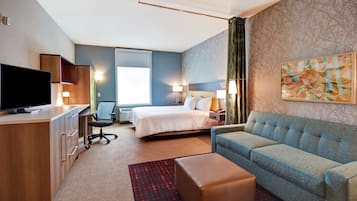 Quarto de hotel com cama, sofá, escrivaninha com televisão e um quadro na parede.