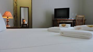 Quarto de hotel com uma cama bem arrumada, uma televisão em um suporte de madeira e um abajur em uma mesinha de cabeceira.