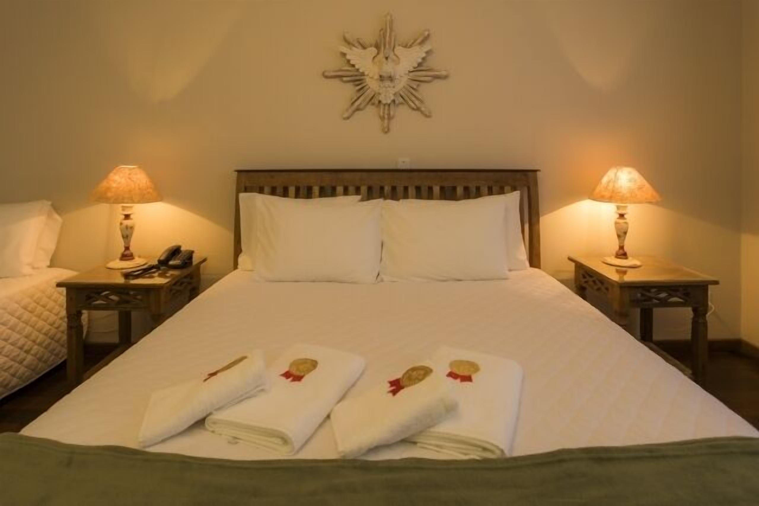 Quarto de hotel com uma cama grande, duas mesas de cabeceira com abajures, e um objeto decorativo acima da cama.