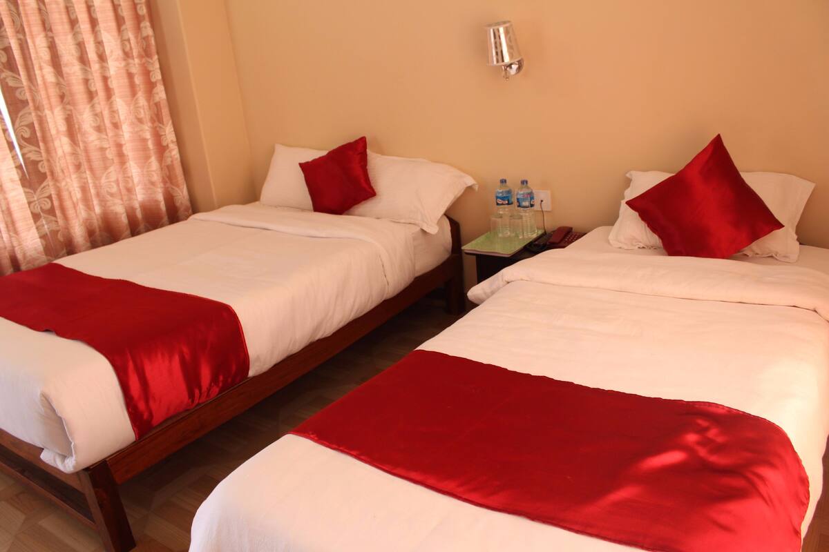 Duas camas de solteiro com almofadas vermelhas e roupa de cama branca, uma mesa de cabeceira com garrafas de água e uma luminária na parede.