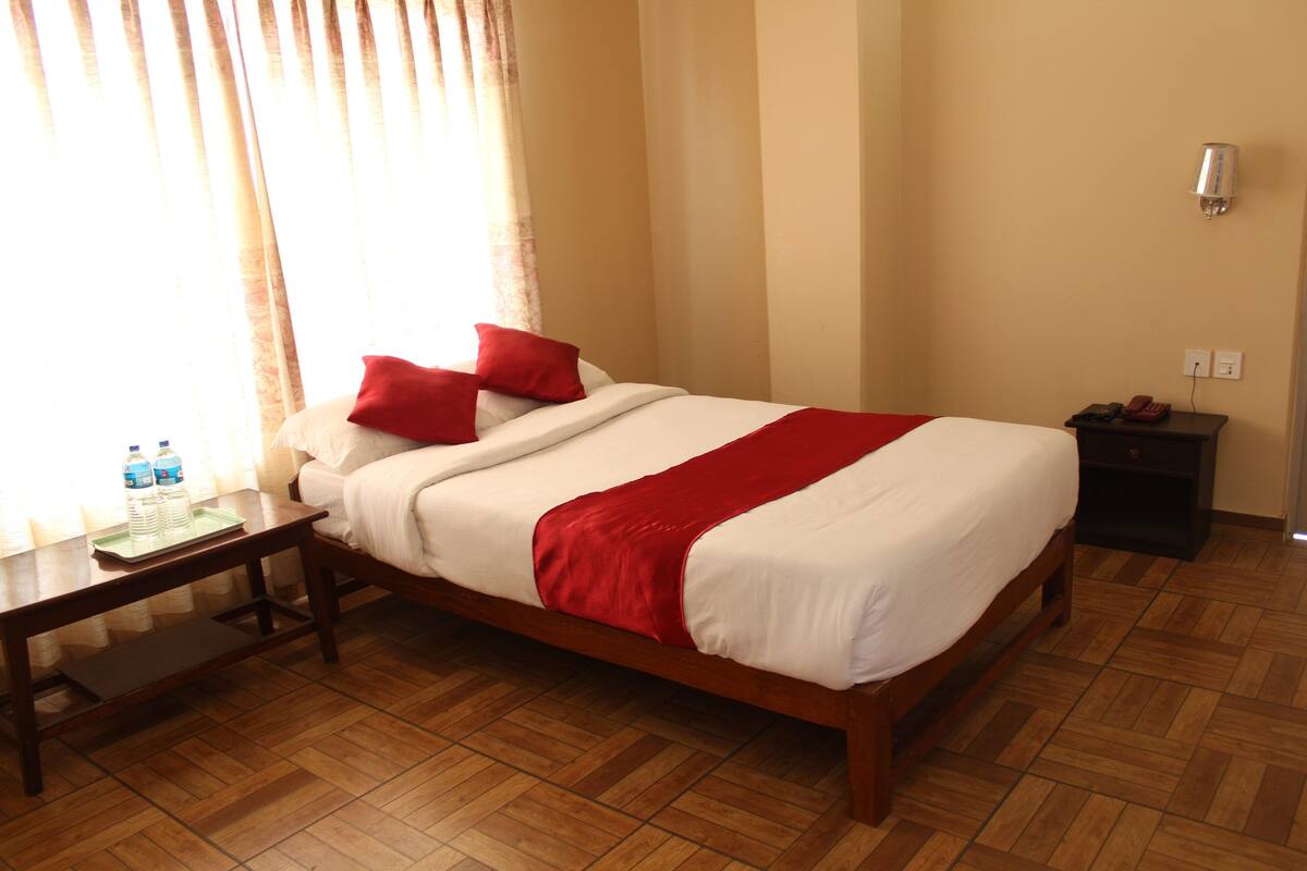 Uma cama bem arrumada com roupa de cama branca e vermelha, uma mesa de cabeceira de madeira com abajur e piso de madeira.