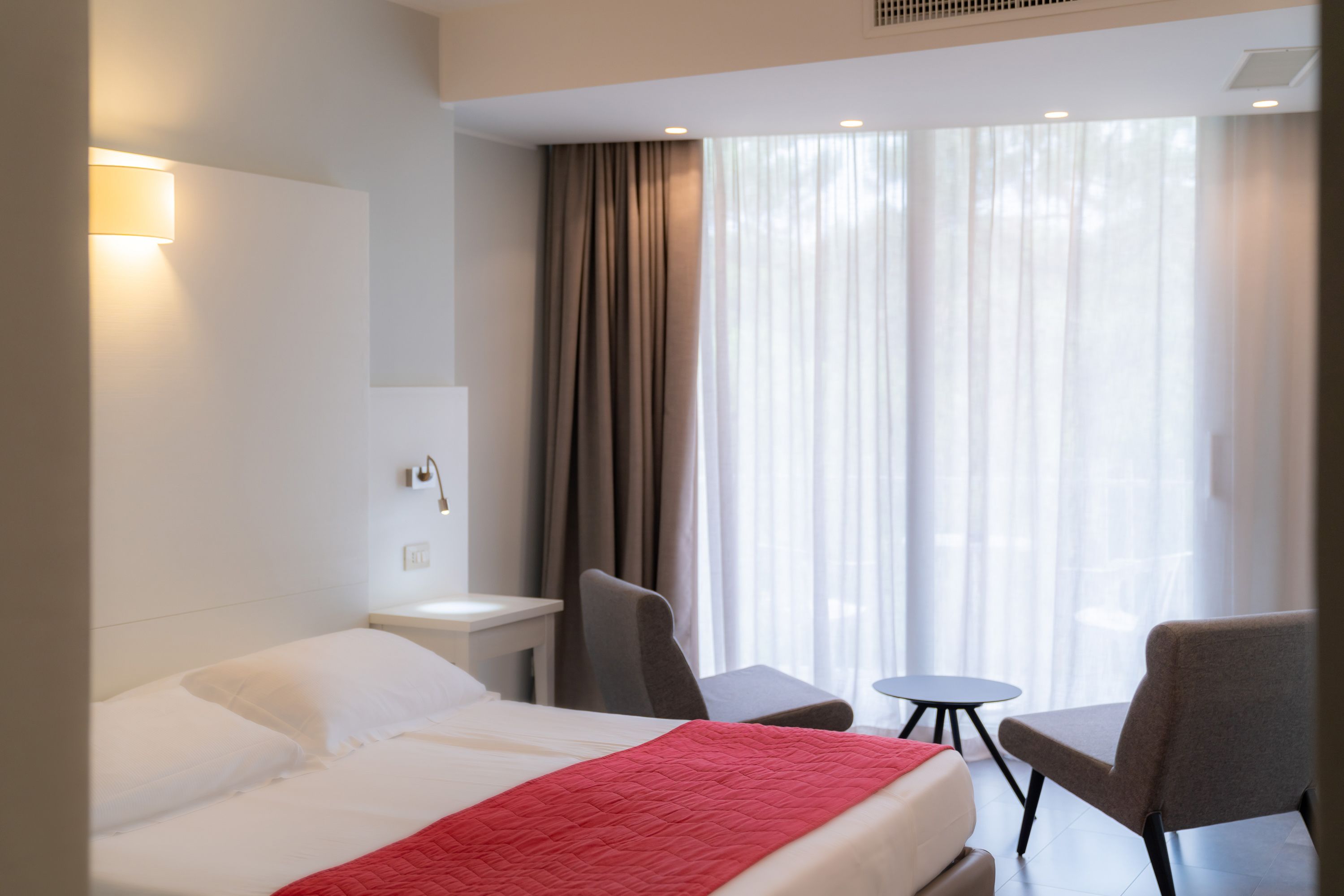 Quarto de hotel com cama, escrivaninha, duas cadeiras, mesa pequena e uma janela grande com cortinas.