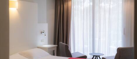 Quarto de hotel com cama, escrivaninha, duas cadeiras, mesa pequena e uma janela grande com cortinas.