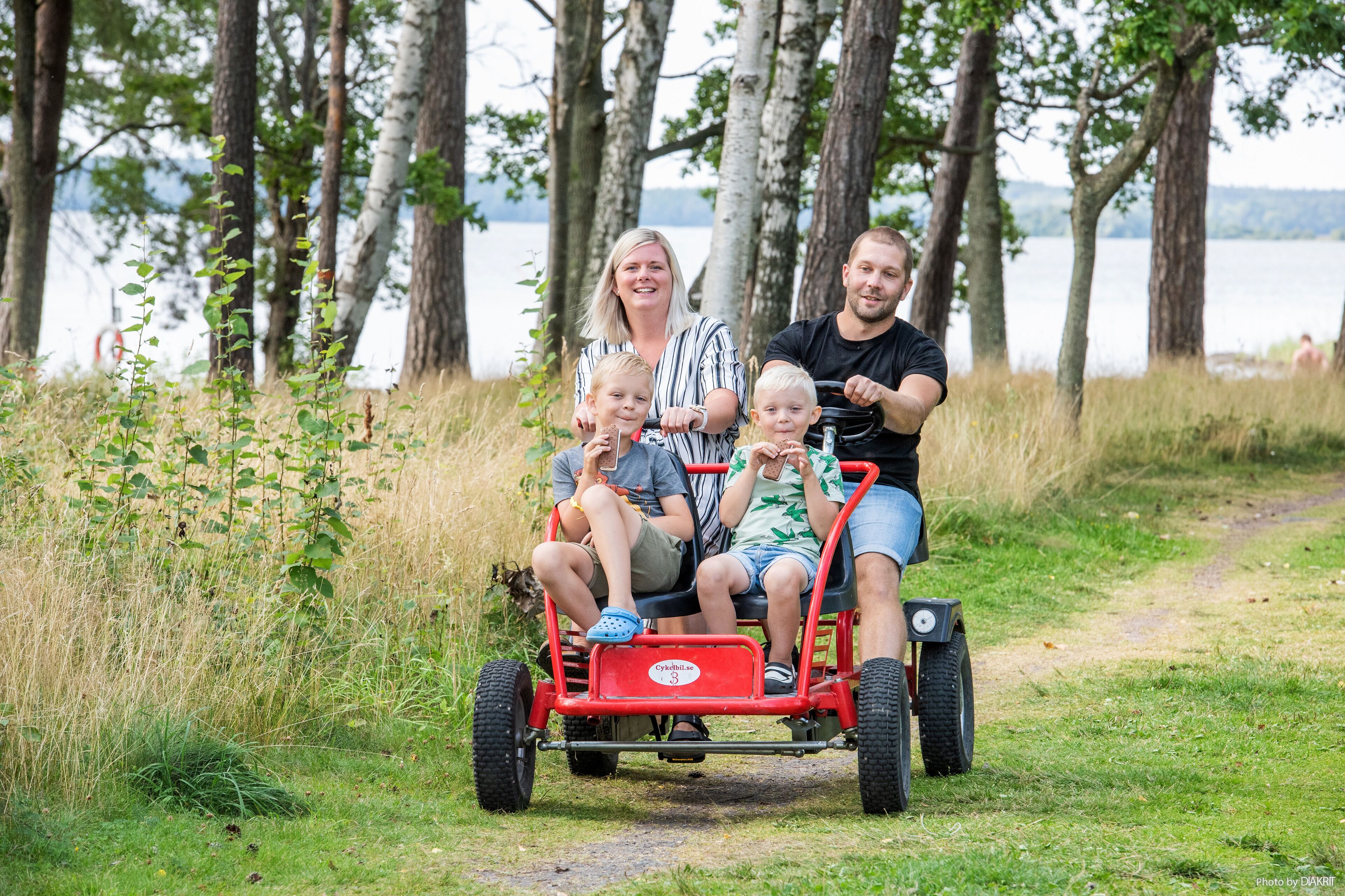 Uma família em um carrinho vermelho perto de um lago, com árvores ao fundo.