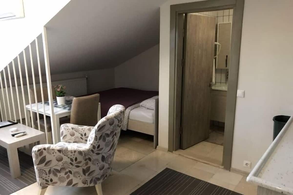 Quarto pequeno e moderno com uma cama, uma cadeira, uma mesa e vista para um banheiro por uma porta aberta.