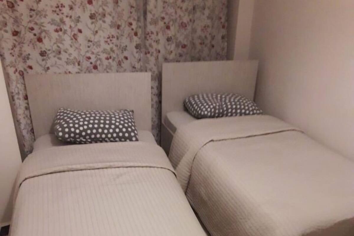 Quarto com duas camas de solteiro, cada uma com almofada estampada e cabeceira lisa. O quarto possui piso de azulejos e cortinas florais na janela.