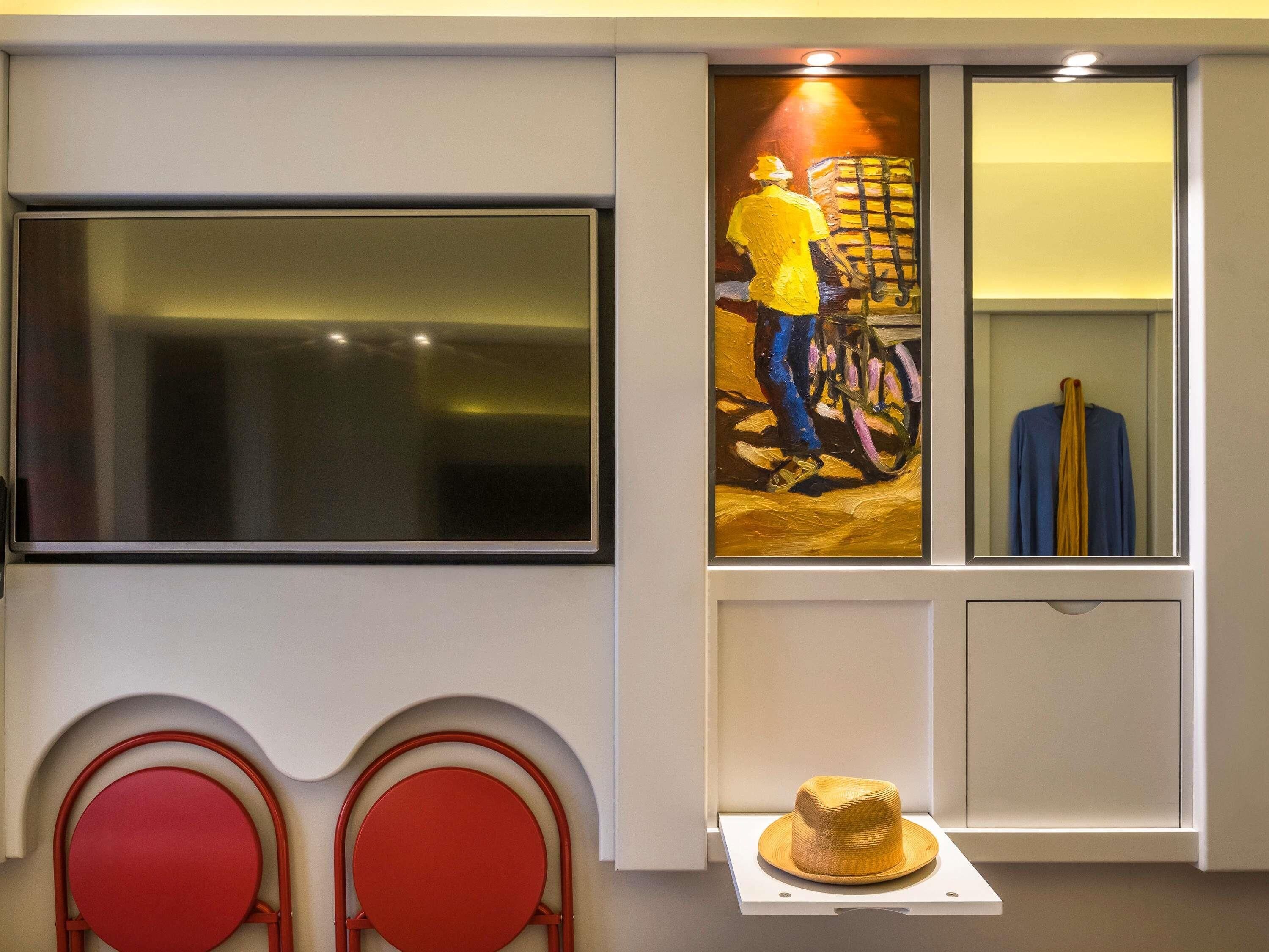 Quarto moderno com uma televisão de tela plana grande fixada na parede, uma mesinha com um chapéu e duas cadeiras vermelhas.