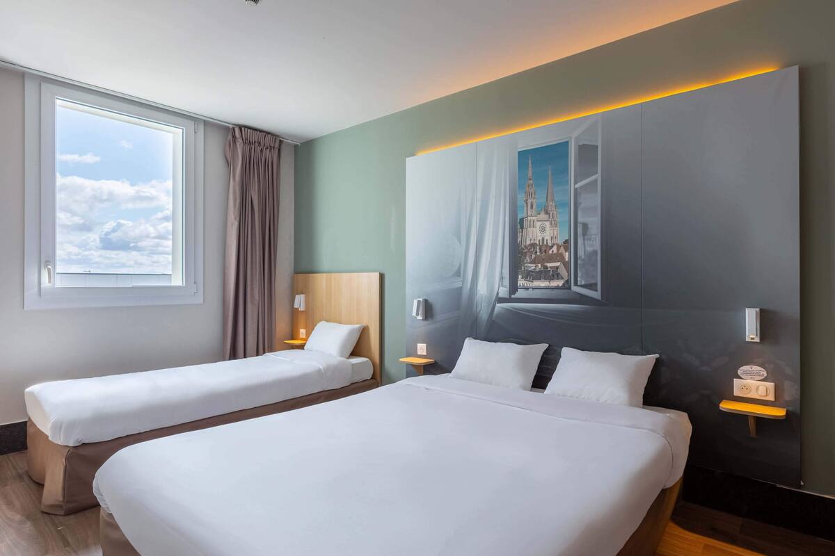 Quarto de hotel com duas camas, uma janela grande com vista do céu, um quadro na parede e uma mesa de cabeceira com abajur.