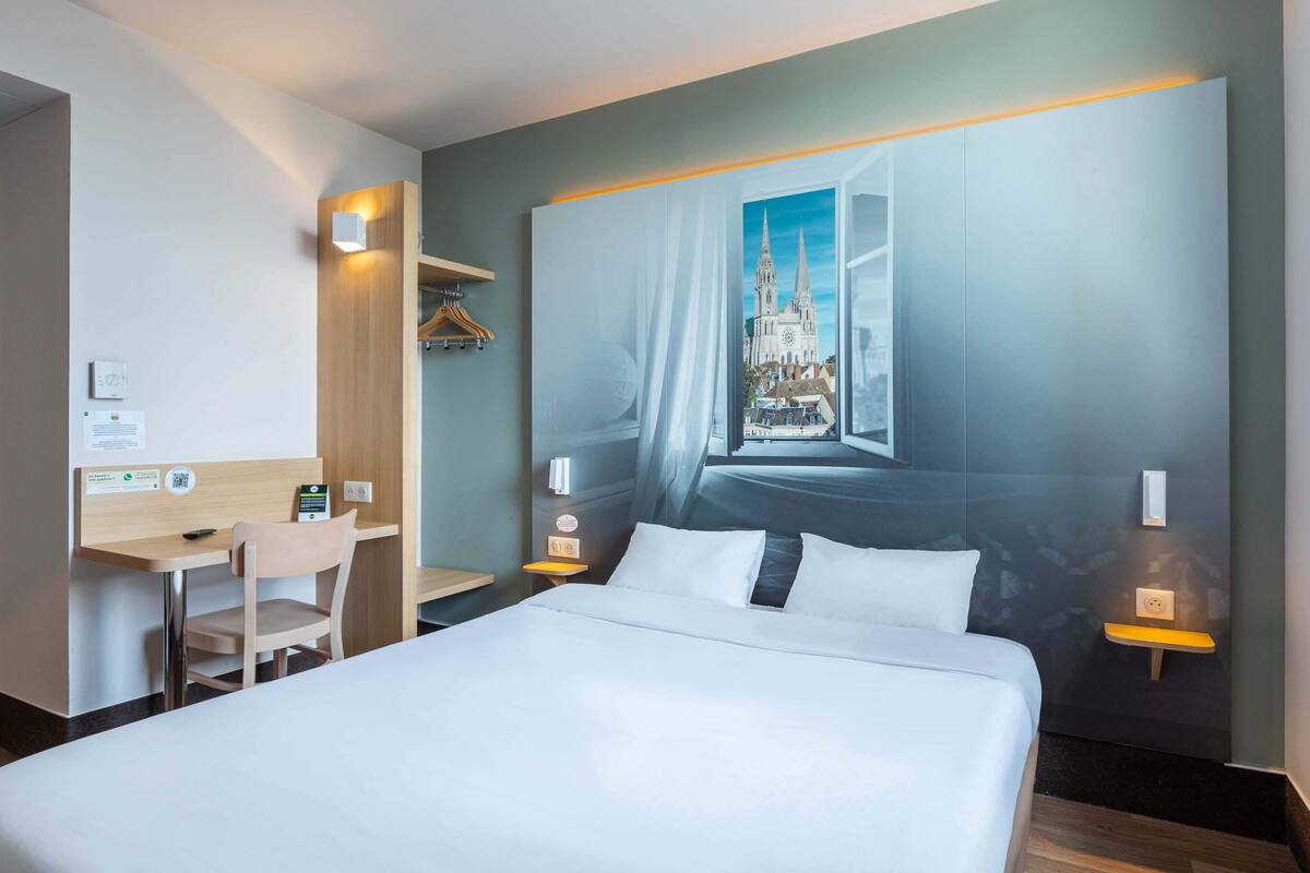 Quarto de hotel com cama, escrivaninha, cadeira e pintura de uma catedral na parede.