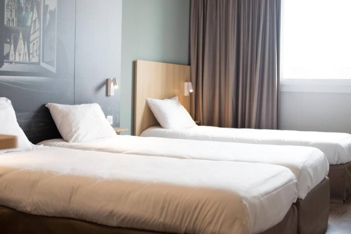 Quarto de hotel com duas camas de solteiro, cabeceira de madeira, mesa de cabeceira e uma grande janela com cortinas.
