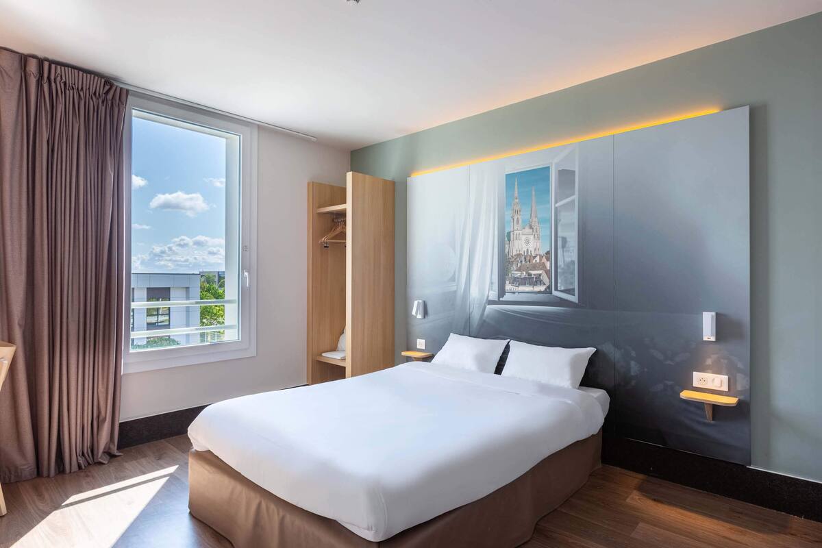 Quarto de hotel moderno com uma cama grande, um guarda-roupa, uma mesa de cabeceira e vista para um edifício pela janela.