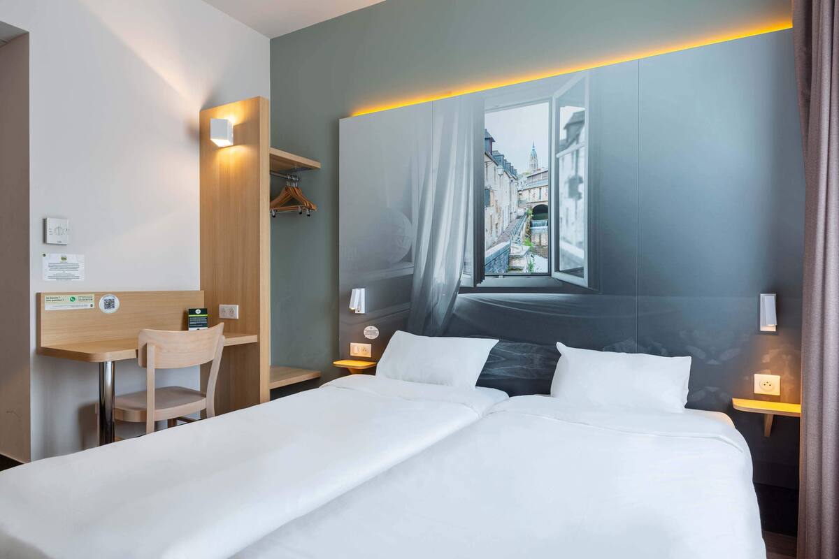 Quarto de hotel com uma cama grande, uma escrivaninha com cadeira, mesa de cabeceira e vista para a cidade pela janela.
