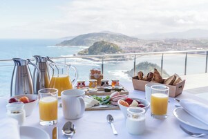 Mesa de café da manhã com diversos alimentos e bebidas, com vista para a paisagem costeira.