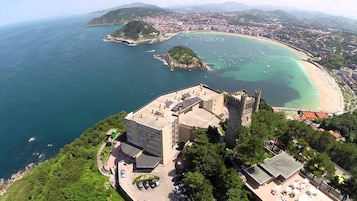 Vista aérea de uma cidade costeira com uma estrutura imponente semelhante a um castelo, uma praia e um porto de iates.