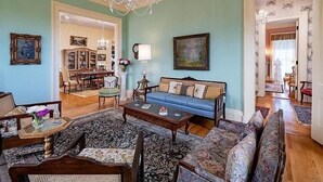 Uma sala de estar espaçosa com um candelabro, um sofá azul, tapetes estampados e um espaço de jantar ao fundo.