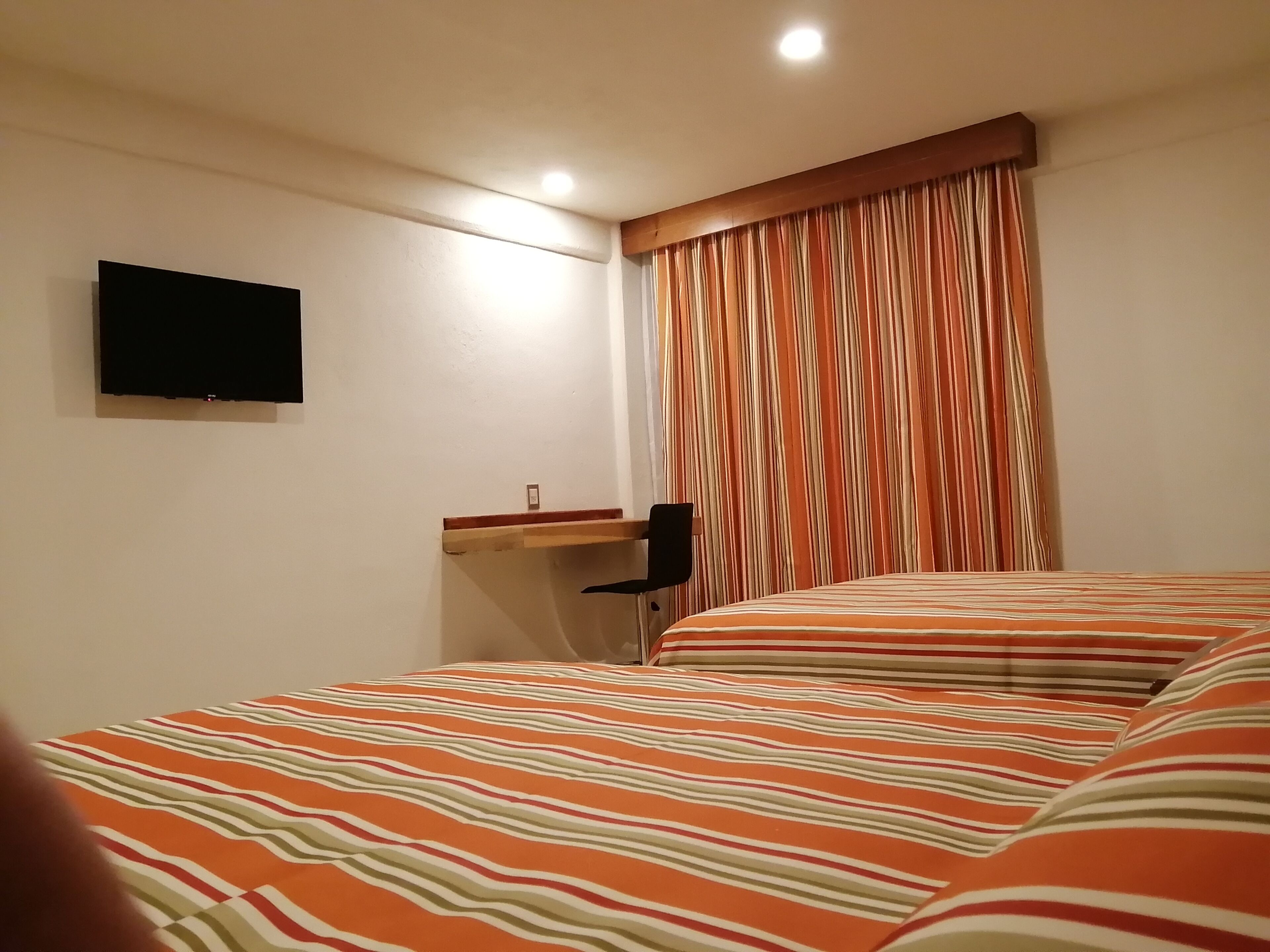 Quarto de hotel com duas camas, televisão fixada na parede, escrivaninha e janela com cortinas listradas.