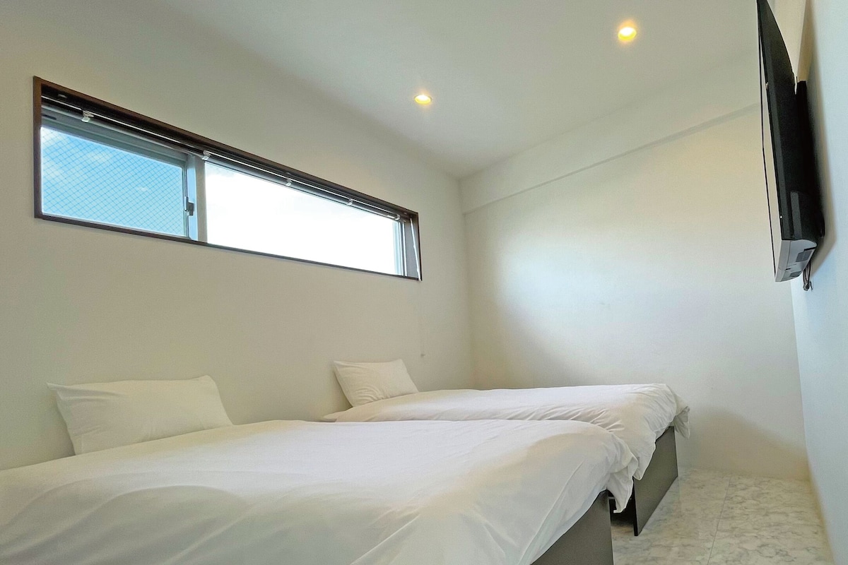 Quarto pequeno e limpo, com duas camas, uma janela e uma TV na parede.