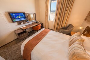 Quarto de hotel com cama, escrivaninha com computador, cadeira, televisão fixada na parede e janela com cortinas.