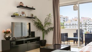 Uma sala de estar moderna com sofá, televisão, mesa de centro, planta em vaso e varanda com mobiliário para exterior.