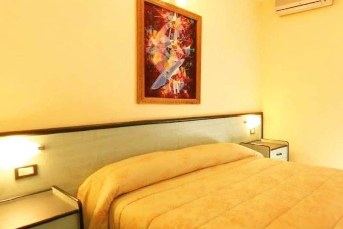 Quarto de hotel com cama, mesas de cabeceira, um quadro na parede e um aparelho de ar condicionado.