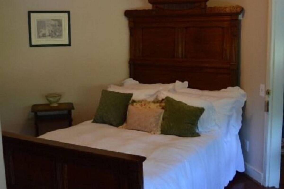 Quarto com cabeceira de madeira, cama com roupa de cama branca e uma pequena mesa de cabeceira com abajur.