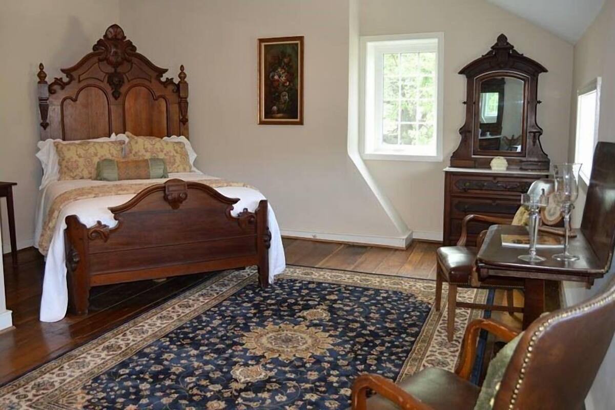 Quarto com cama de madeira grande, penteadeira clássica, espelho e janela com luz natural.