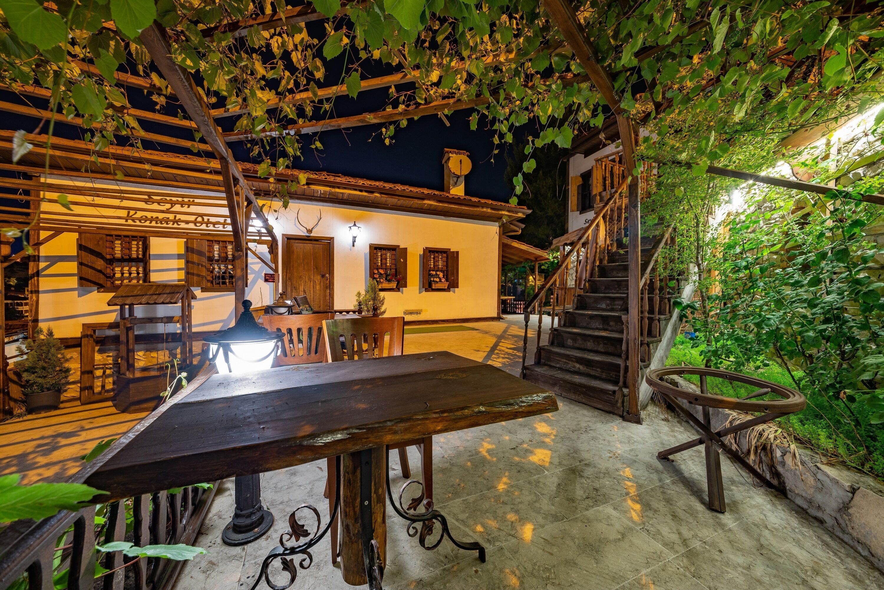 Uma área de estar ao ar livre rústica, com mesa e cadeiras de madeira, cercada por vegetação e uma escadaria que leva a um edifício com uma placa indicando 