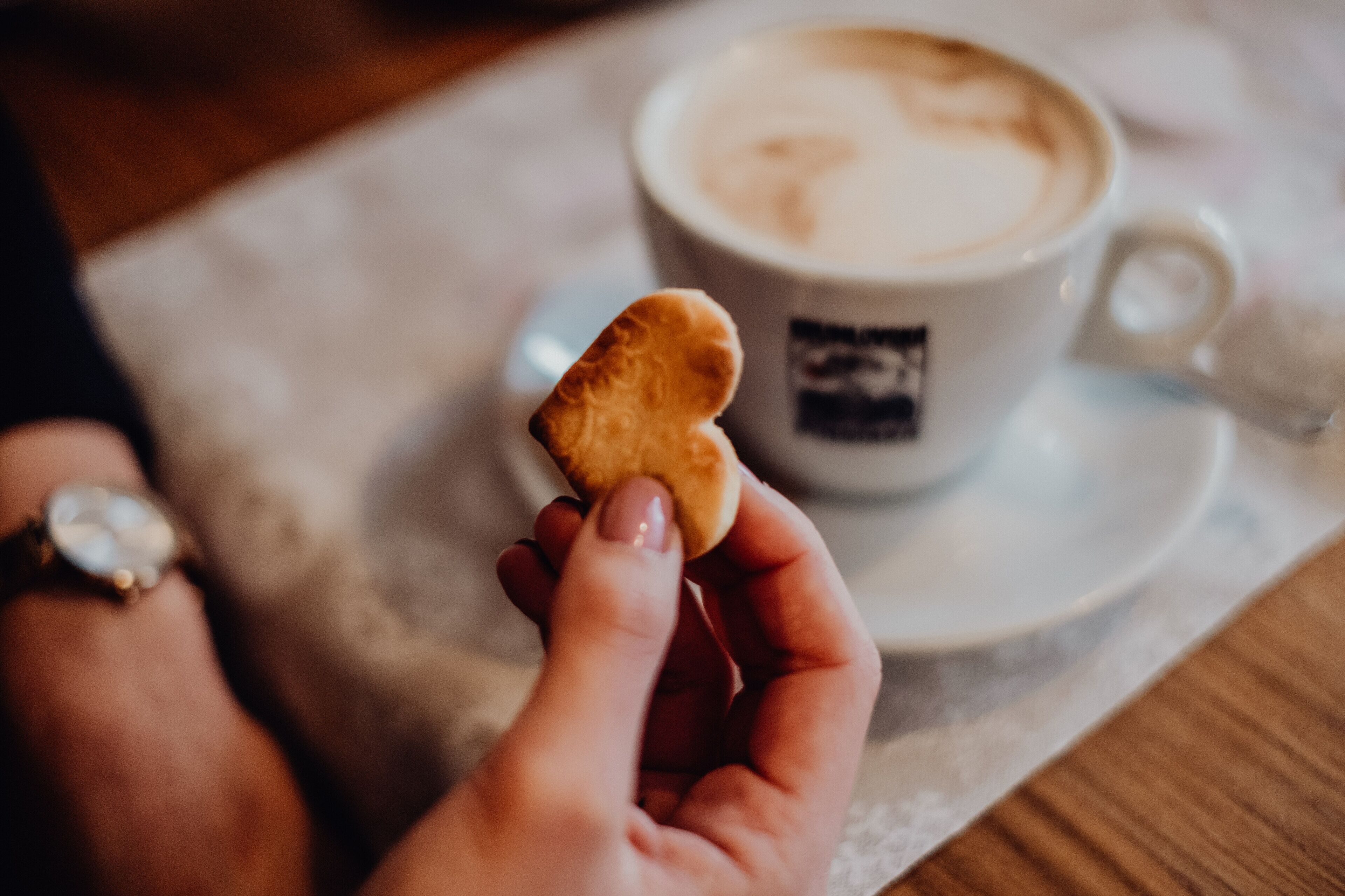Uma mão segurando um objeto pequeno, redondo e marrom-claro, possivelmente um biscoito, em frente a uma xícara de café com um pires branco.