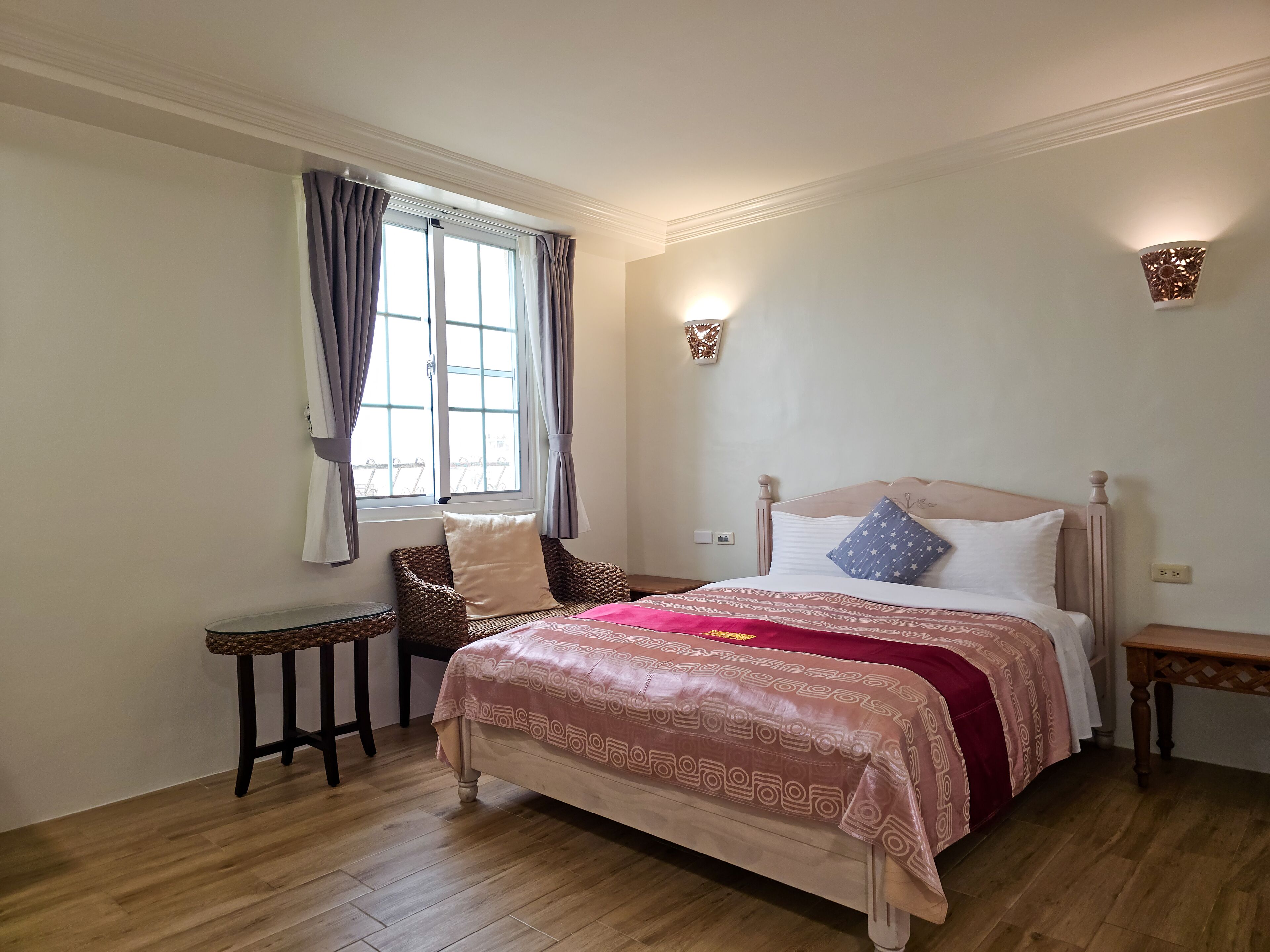 Quarto com cama, uma cadeira, mesa pequena, janela com cortinas e luminária fixada na parede.