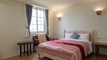 Quarto com cama, uma cadeira, mesa pequena, janela com cortinas e luminária fixada na parede.