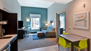 Quarto de hotel moderno com cozinha compacta, área de estar com sofá e cadeiras, mesa de jantar com cadeiras amarelas e uma janela grande com persianas.
