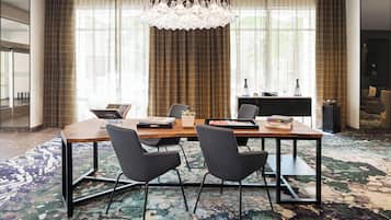 Uma sala de conferências com uma mesa de madeira, quatro cadeiras, um candelabro e uma janela ampla com cortinas.