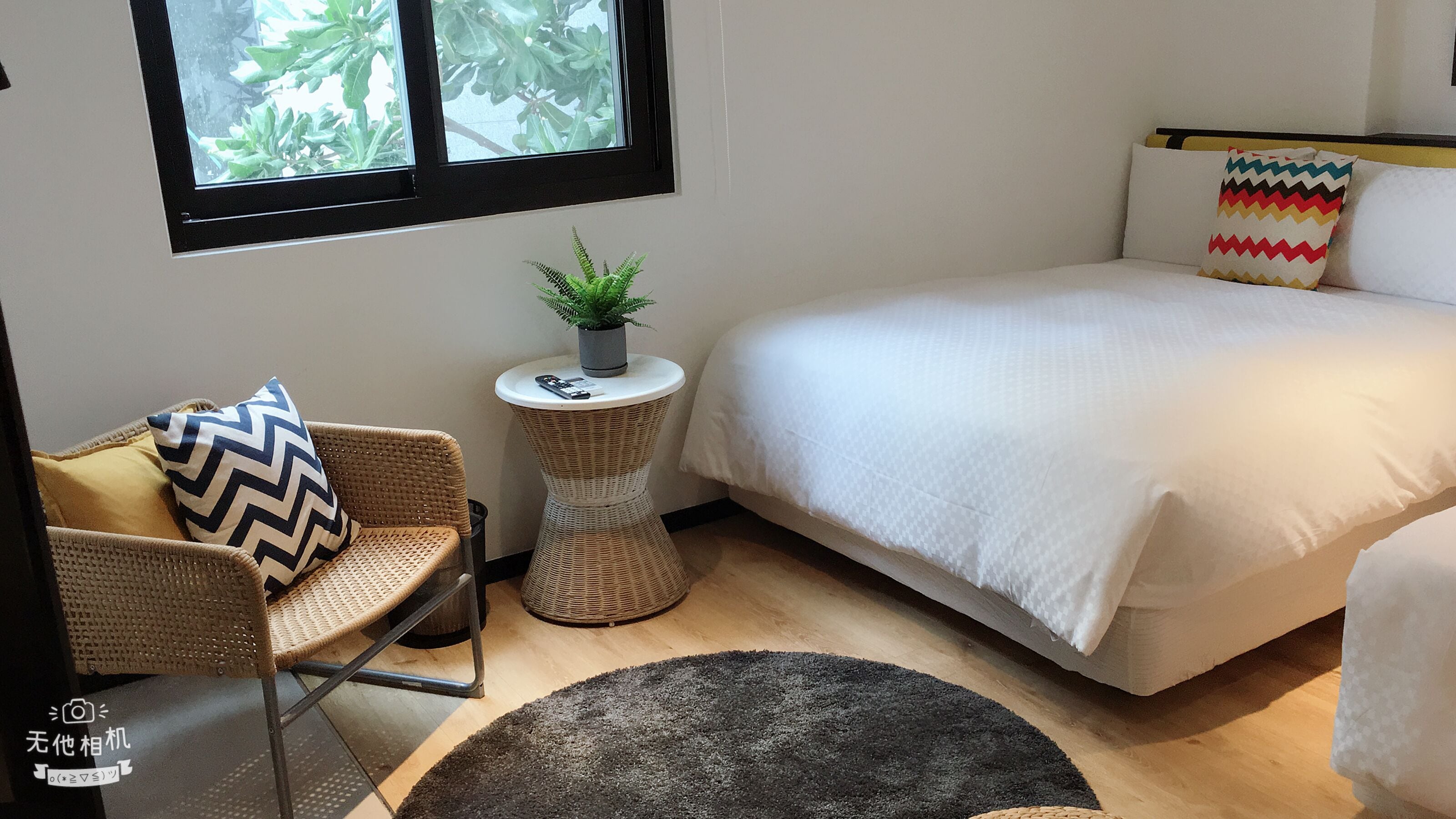 Quarto com uma cama, uma cadeira, uma mesinha com uma planta e uma janela com vista para árvores.