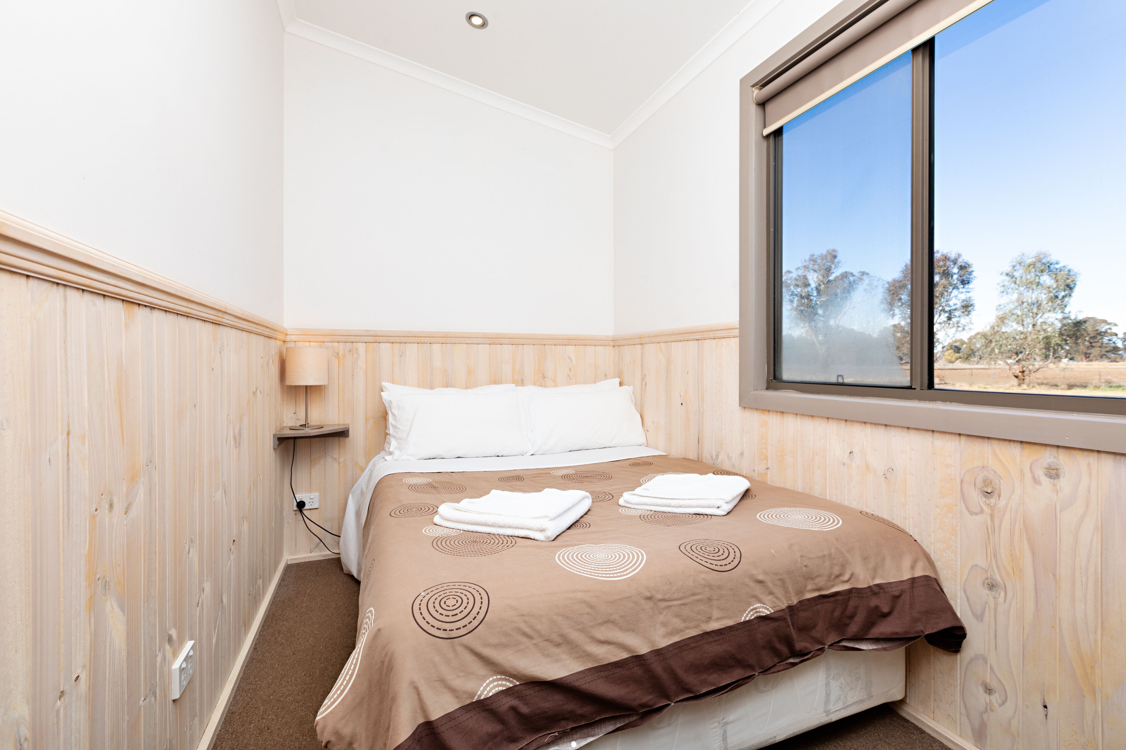 Cama bem arrumada com colcha marrom e branca estampada, cabeceira de madeira e janela com vista para árvores.