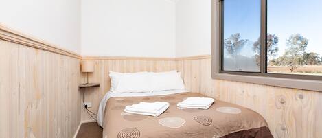 Cama bem arrumada com colcha marrom e branca estampada, cabeceira de madeira e janela com vista para árvores.