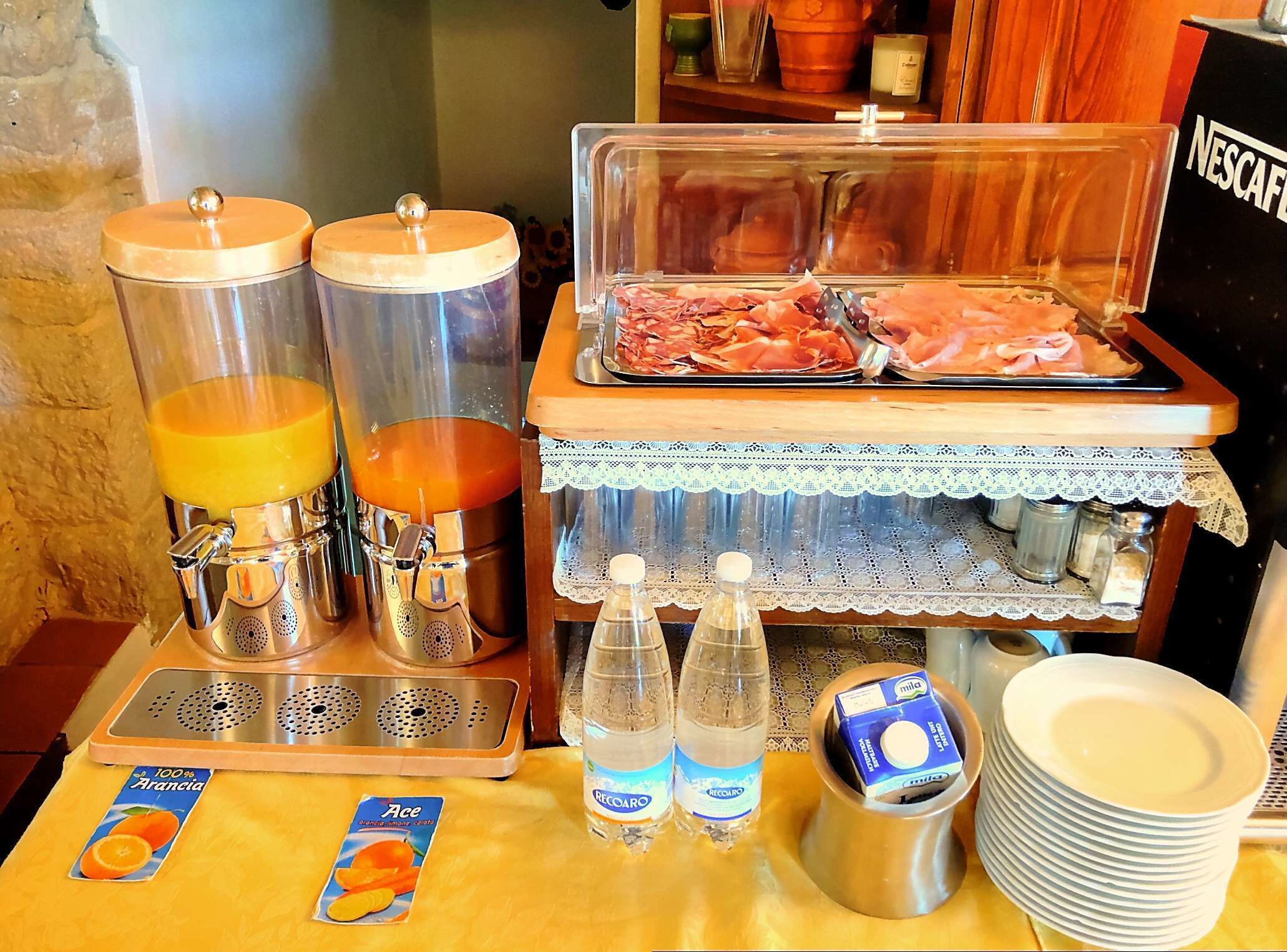 Uma estação de café da manhã com dispensadores de suco, uma máquina de café, um balcão de frios, garrafas de água e uma pilha de pratos.