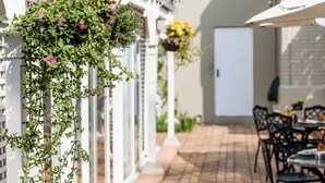 Uma área de pátio com assentos externos, um cesto de flores pendurado e uma porta branca ao fundo.
