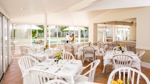 Restaurante com mesas e cadeiras brancas, vista para piscina e uma pessoa ao fundo.