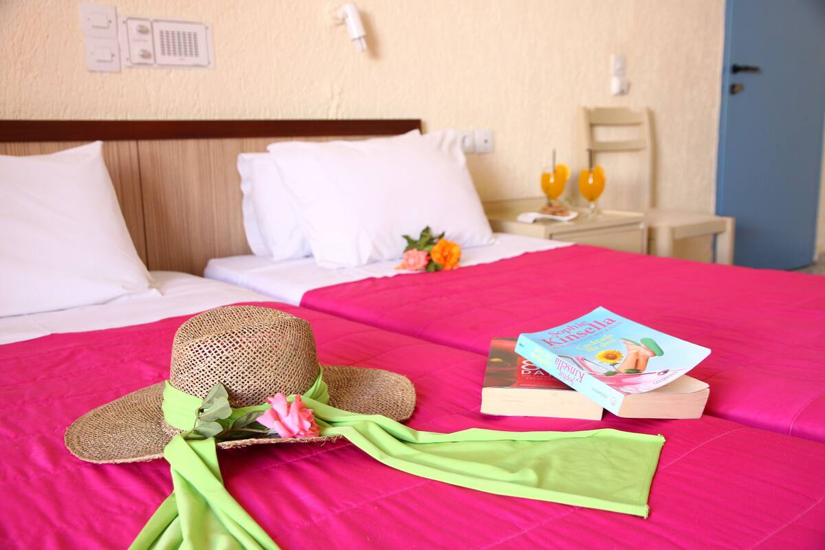 Quarto de hotel com cama, almofadas, um chapéu, um livro e uma mesa de cabeceira com copos.