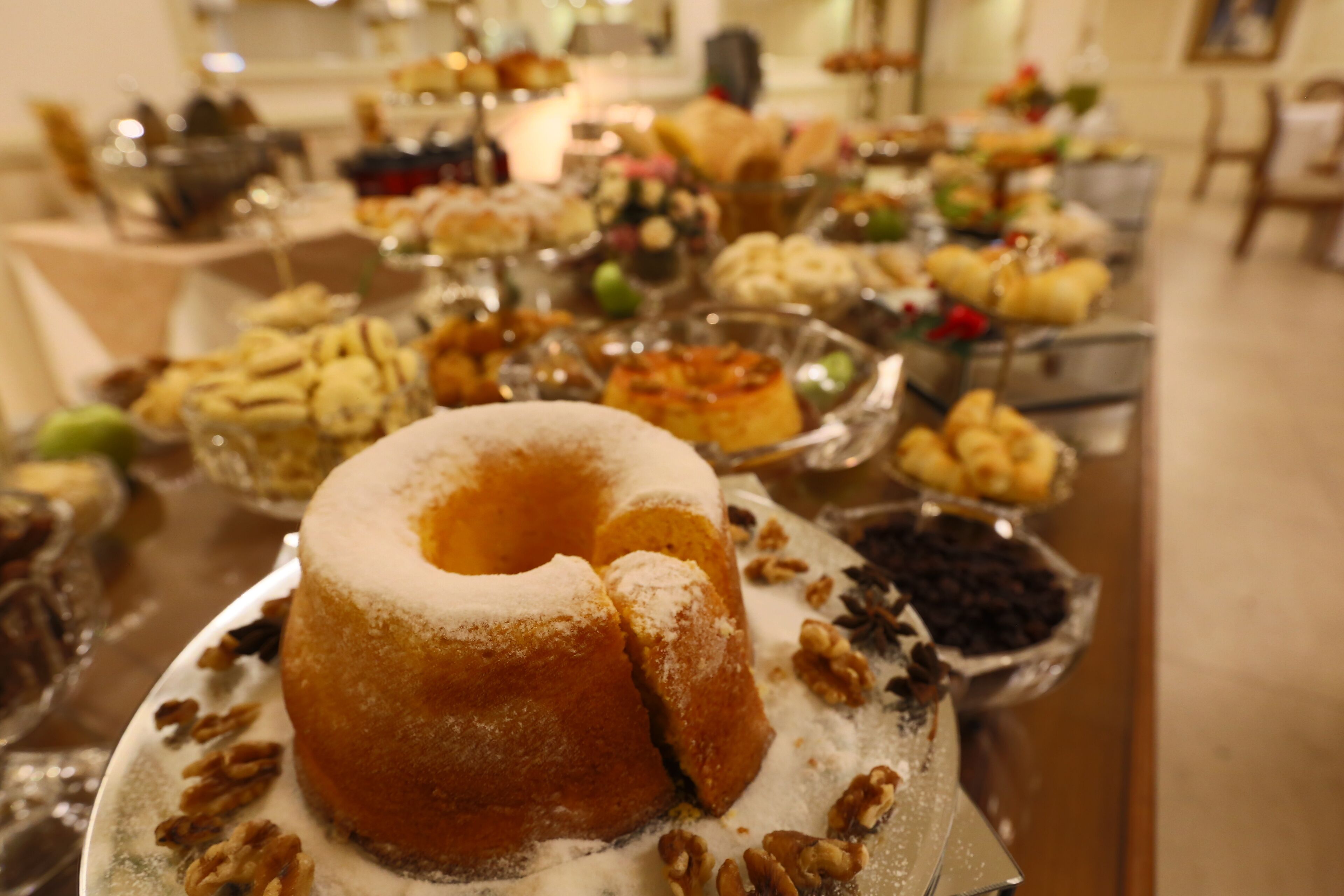 Uma mesa de buffet com diversos pratos, incluindo um bolo bundt central e sobremesas variadas.