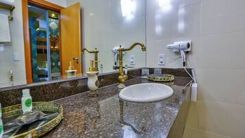 Um banheiro com bancada de mármore, pia branca, torneira dourada e secador de cabelo fixado na parede.