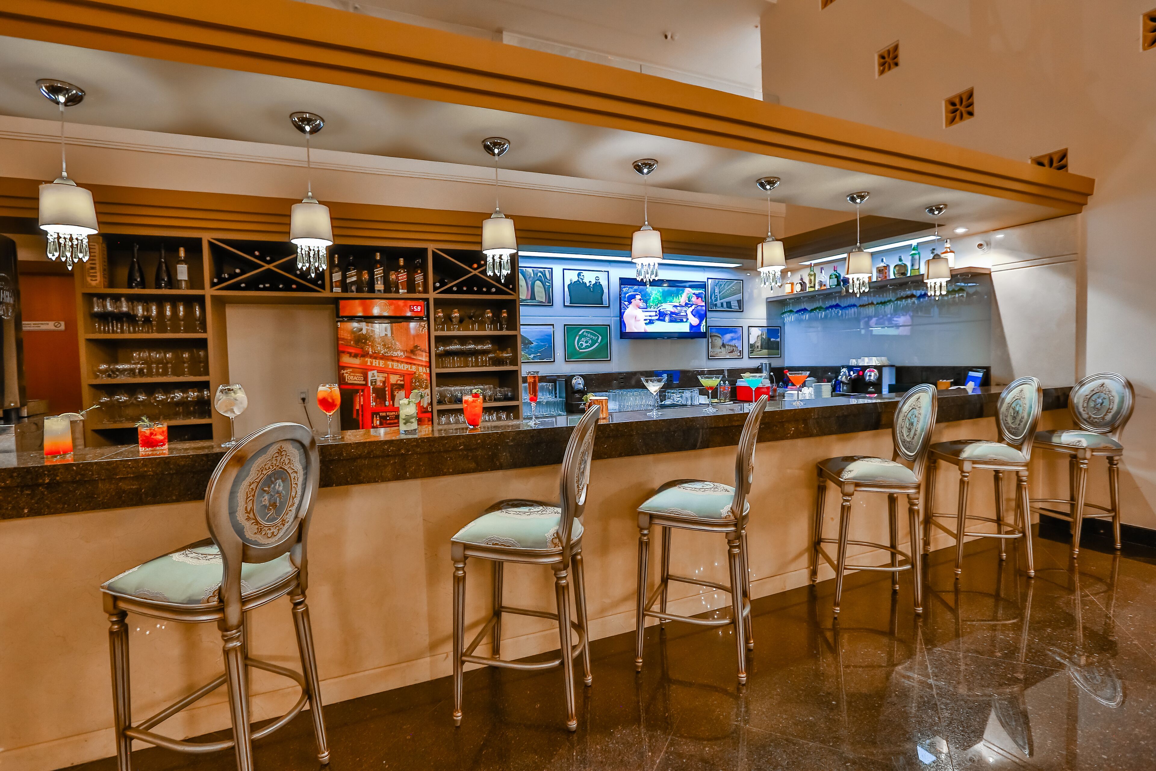 Uma área de bar com vários bancos, prateleiras com garrafas e uma tela de televisão.